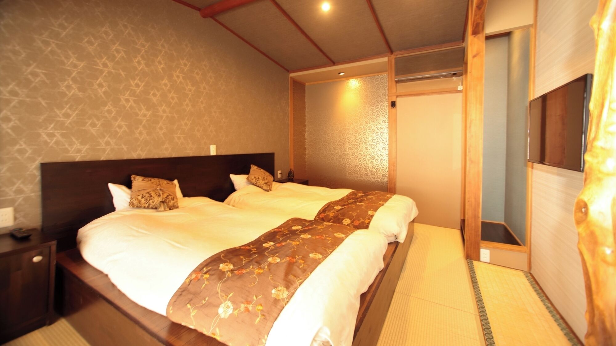 Quarto de hotel com uma cama grande, mesa de cabeceira, cabeceira de madeira e parede com padrão geométrico.