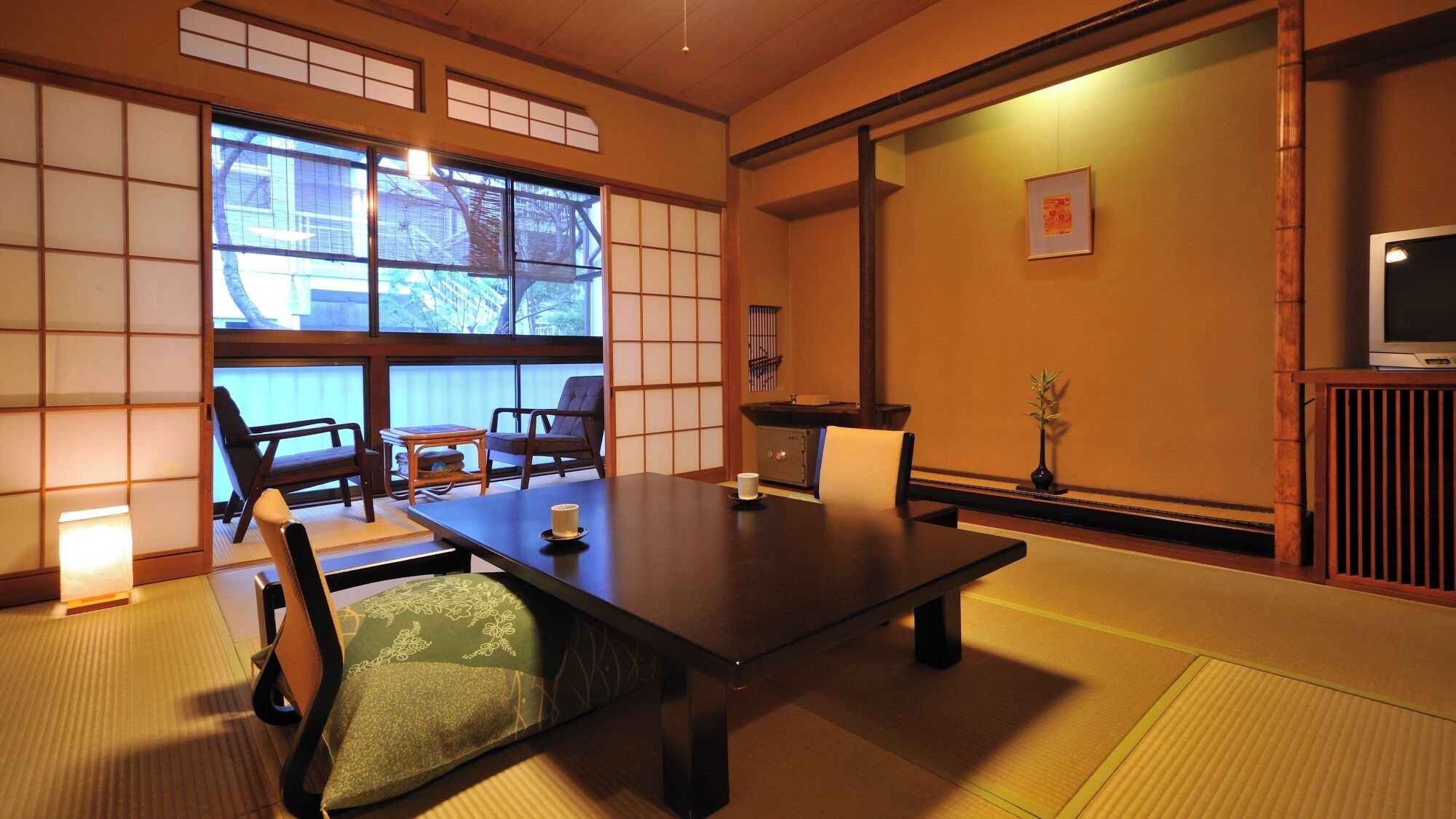 Um quarto japonês tradicional com tatames, uma mesa baixa, cadeiras e uma porta deslizante com vista para o exterior.