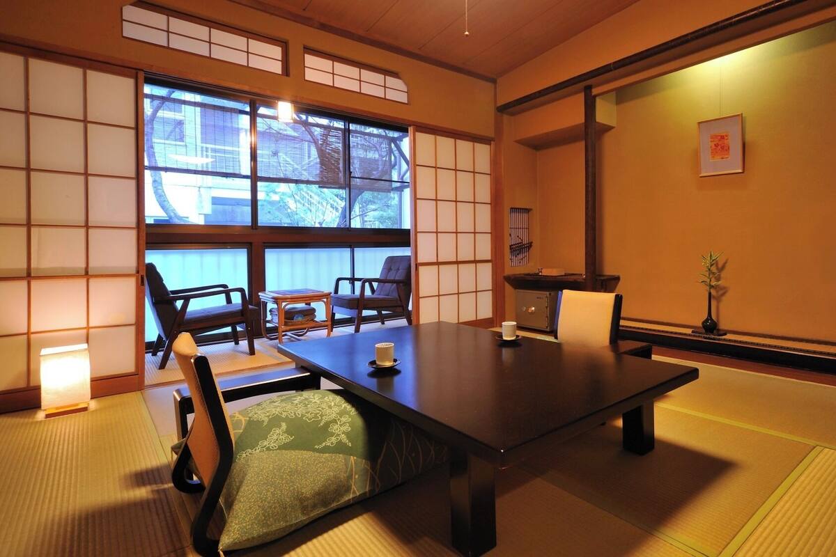 Um quarto japonês tradicional com tatames, uma mesa baixa, cadeiras e uma porta deslizante com vista para o exterior.
