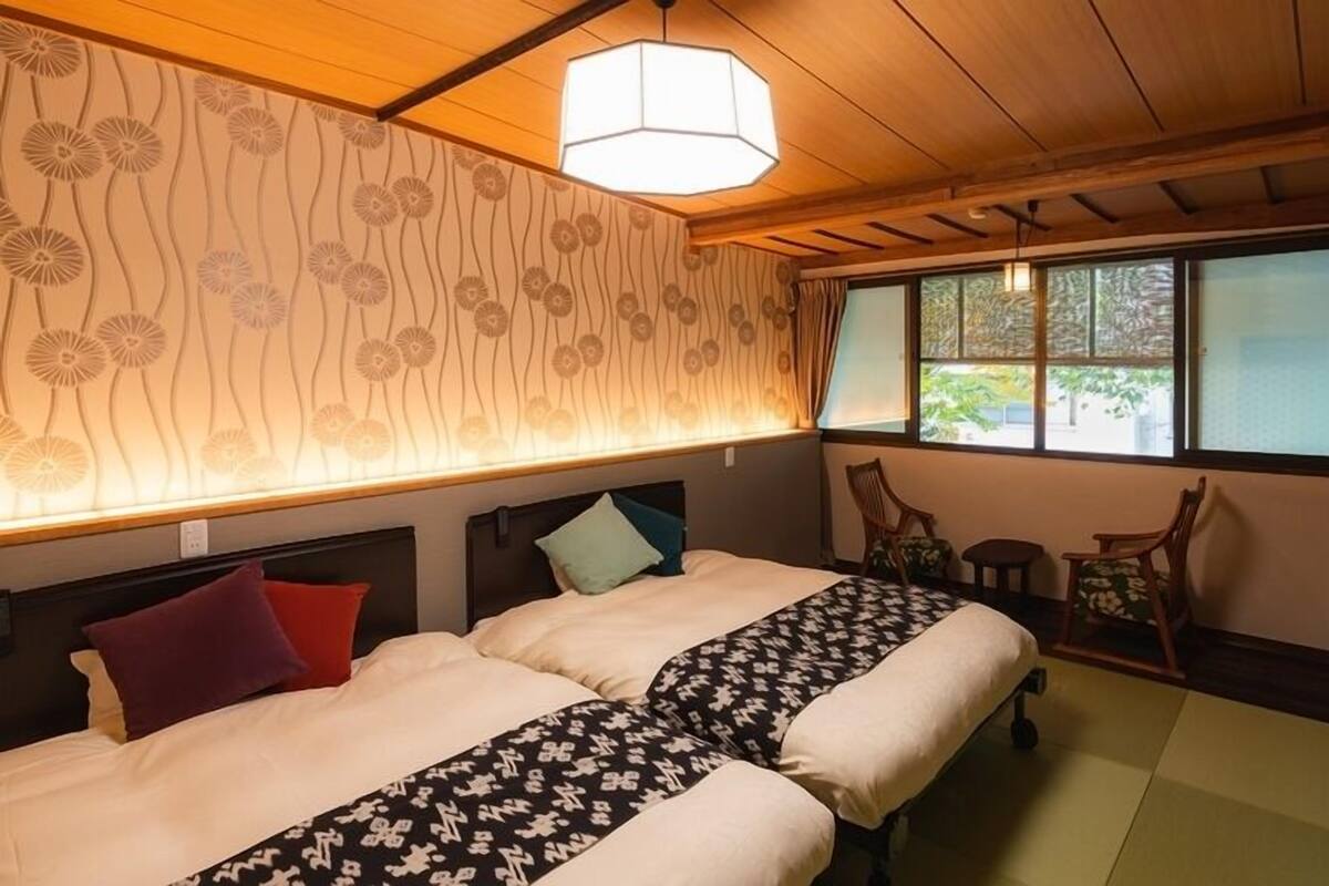 Quarto com duas camas, uma janela com cortinas, uma cadeira e parede estampada.