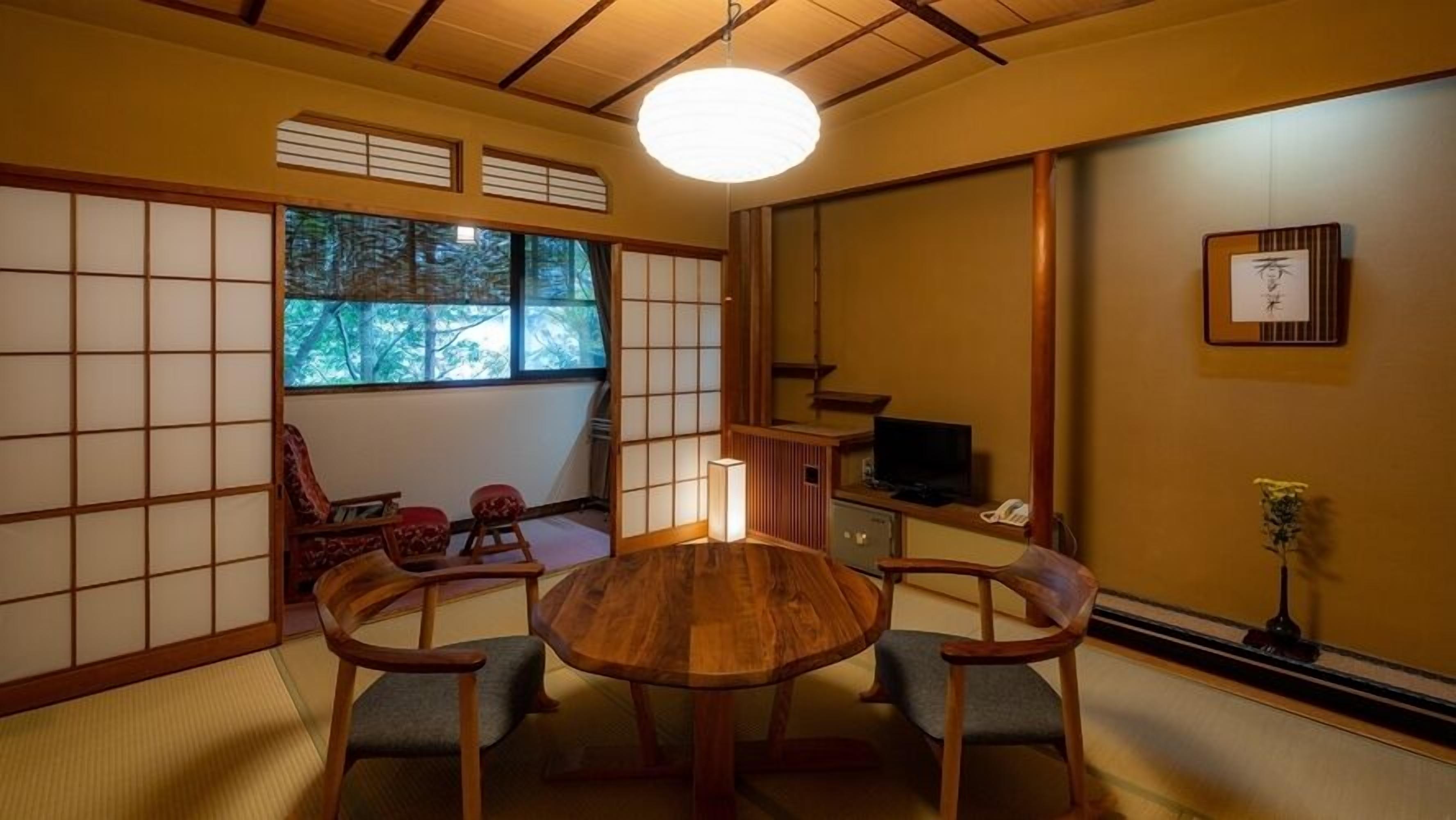 Um quarto japonês tradicional com piso de tatame, uma mesa baixa com quatro cadeiras, uma televisão e uma porta deslizante com vista para árvores.