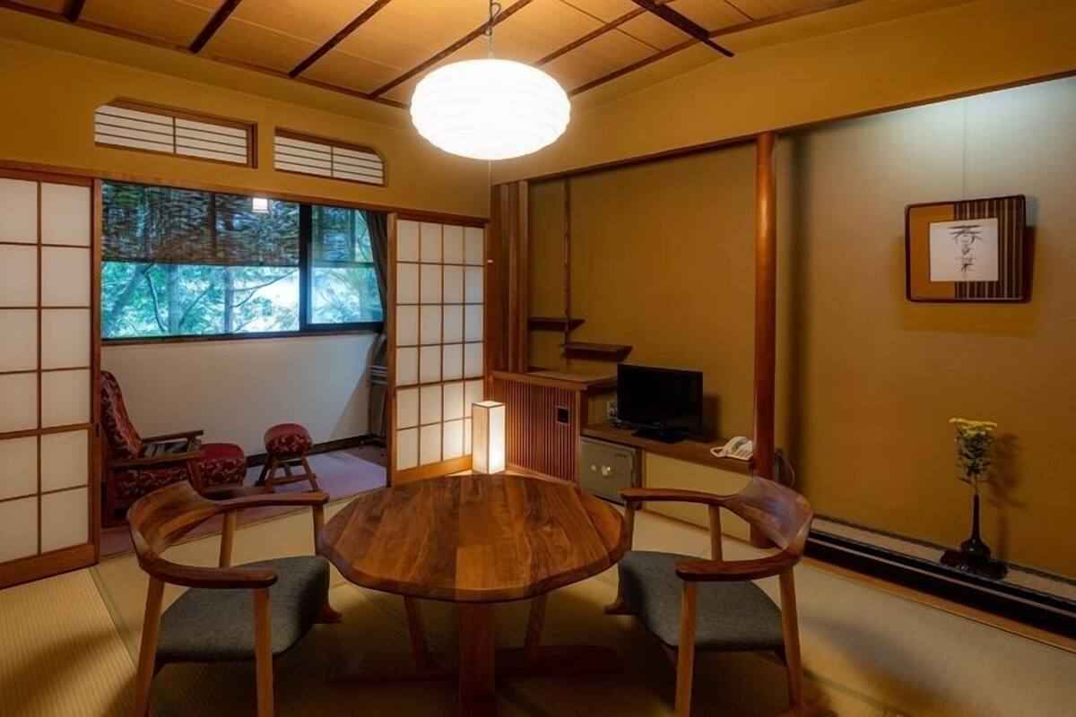 Um quarto japonês tradicional com piso de tatame, uma mesa baixa com quatro cadeiras, uma televisão e uma porta deslizante com vista para árvores.