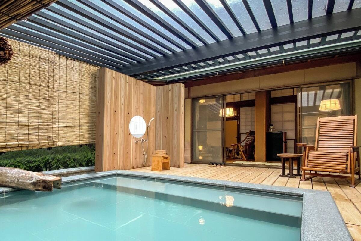 Um deck de madeira com piscina, uma sauna de madeira e uma porta de vidro que dá para um espaço com cadeira e mesa.