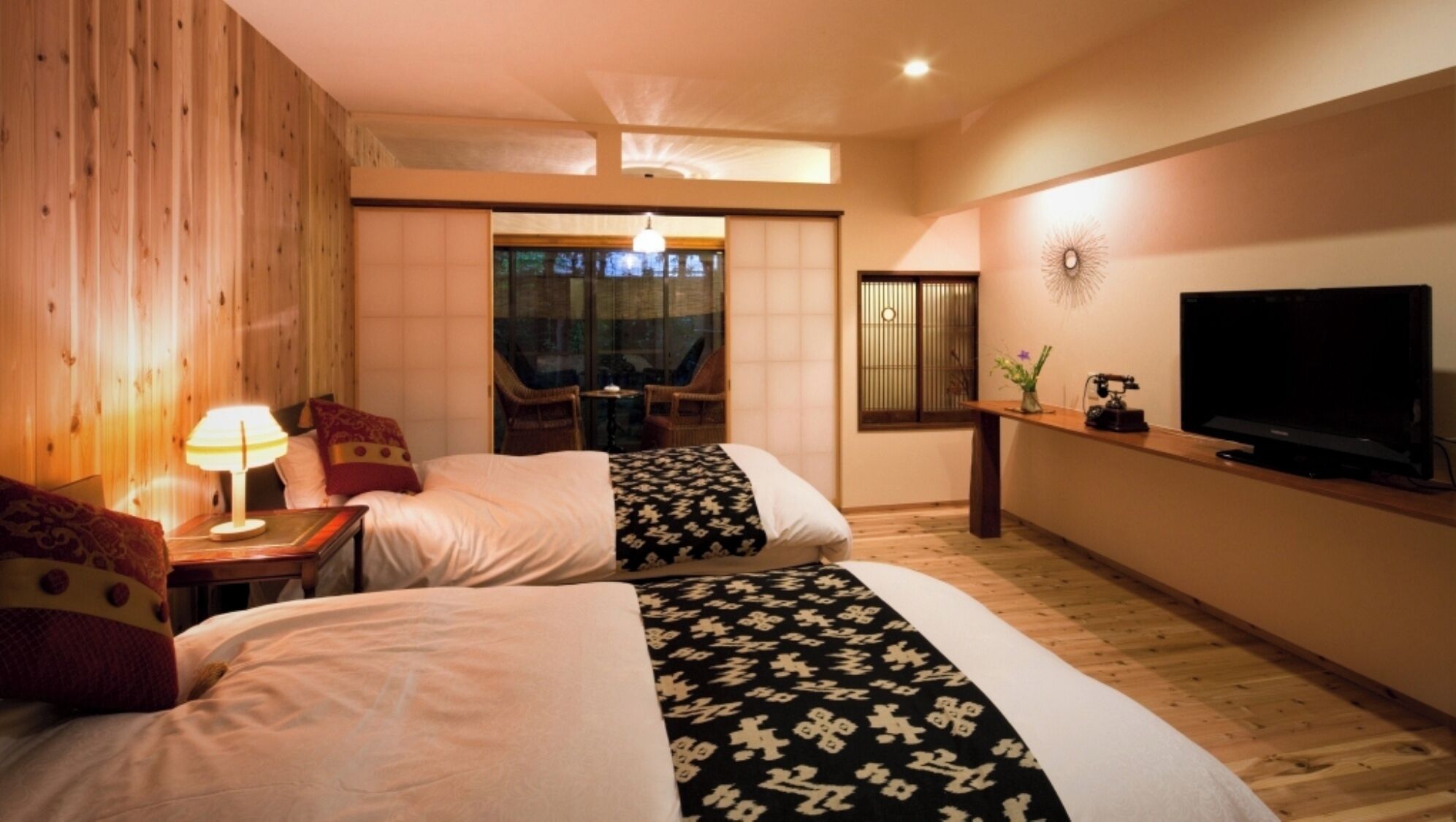 Quarto de hotel com duas camas, televisão, escrivaninha com luminária e vista para o exterior através de uma porta de correr.