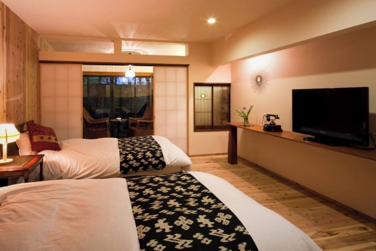 Quarto de hotel com duas camas, televisão, escrivaninha com luminária e vista para o exterior através de uma porta de correr.