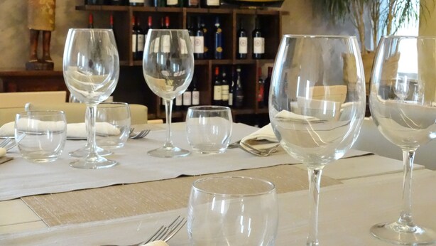 Mesa posta com taças de vinho vazias e uma garrafa de vinho, com um cenário de garrafas de vinho e um barco à vela decorativo.