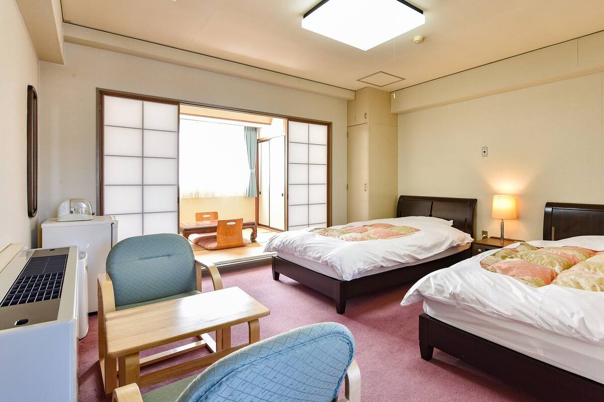 Quarto de hotel com duas camas, uma escrivaninha, uma cadeira, um laptop e janela com vista.