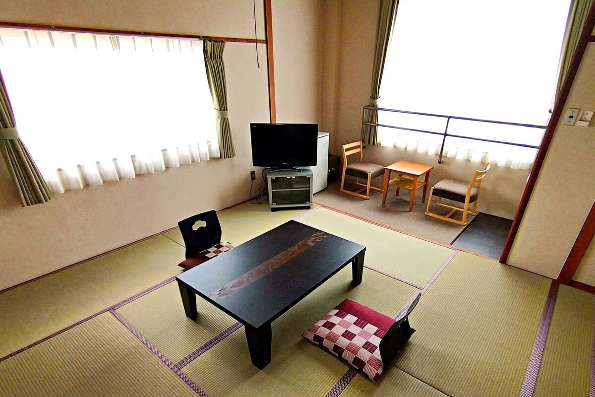 Quarto japonês tradicional com tatames, uma mesa baixa, uma televisão e duas cadeiras.