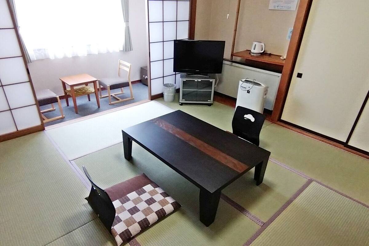 Um quarto japonês tradicional com tatames, uma mesa baixa com almofada, uma televisão e uma pequena cozinha compacta.