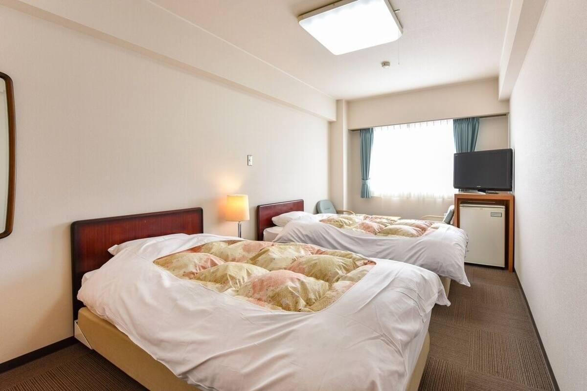 Quarto de hotel com duas camas, uma mesa de trabalho com televisão e uma janela com cortinas.
