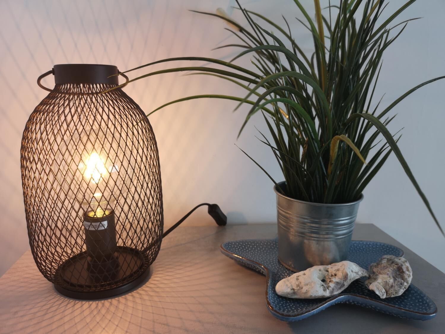 Um candeeiro de metal com uma vela no seu interior, uma planta em vaso e duas pedras decorativas sobre uma bandeja azul.