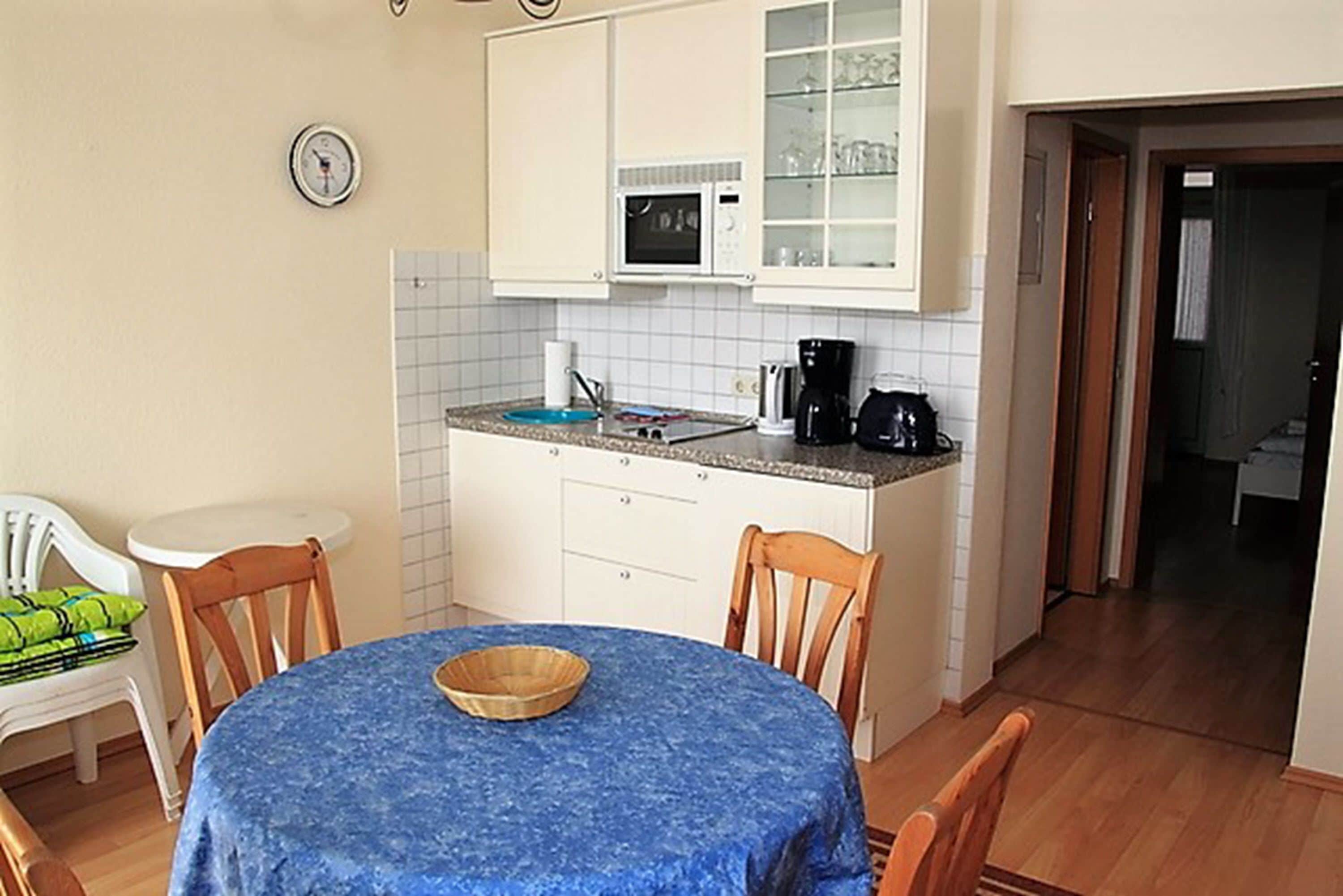 Cozinha compacta com armários brancos, micro-ondas e máquina de café. Há uma mesa de jantar com toalha de mesa azul e uma cadeira de madeira.