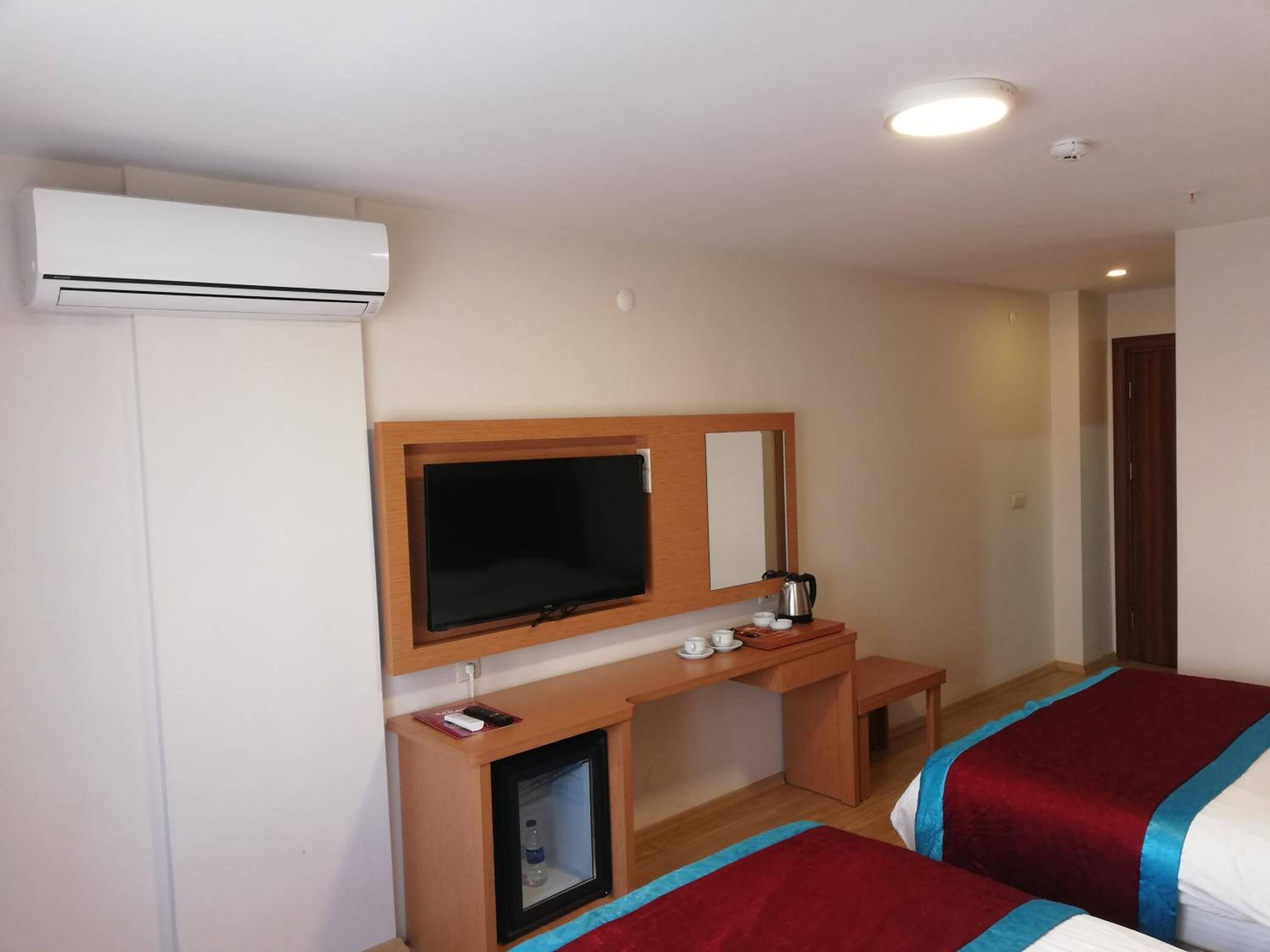 Quarto de hotel com televisão presa a uma escrivaninha de madeira, minibar e duas camas com roupa de cama vermelha e azul.