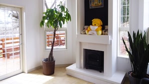 Uma sala de estar com lareira, um vaso de planta e uma janela com vista para um deck de madeira.