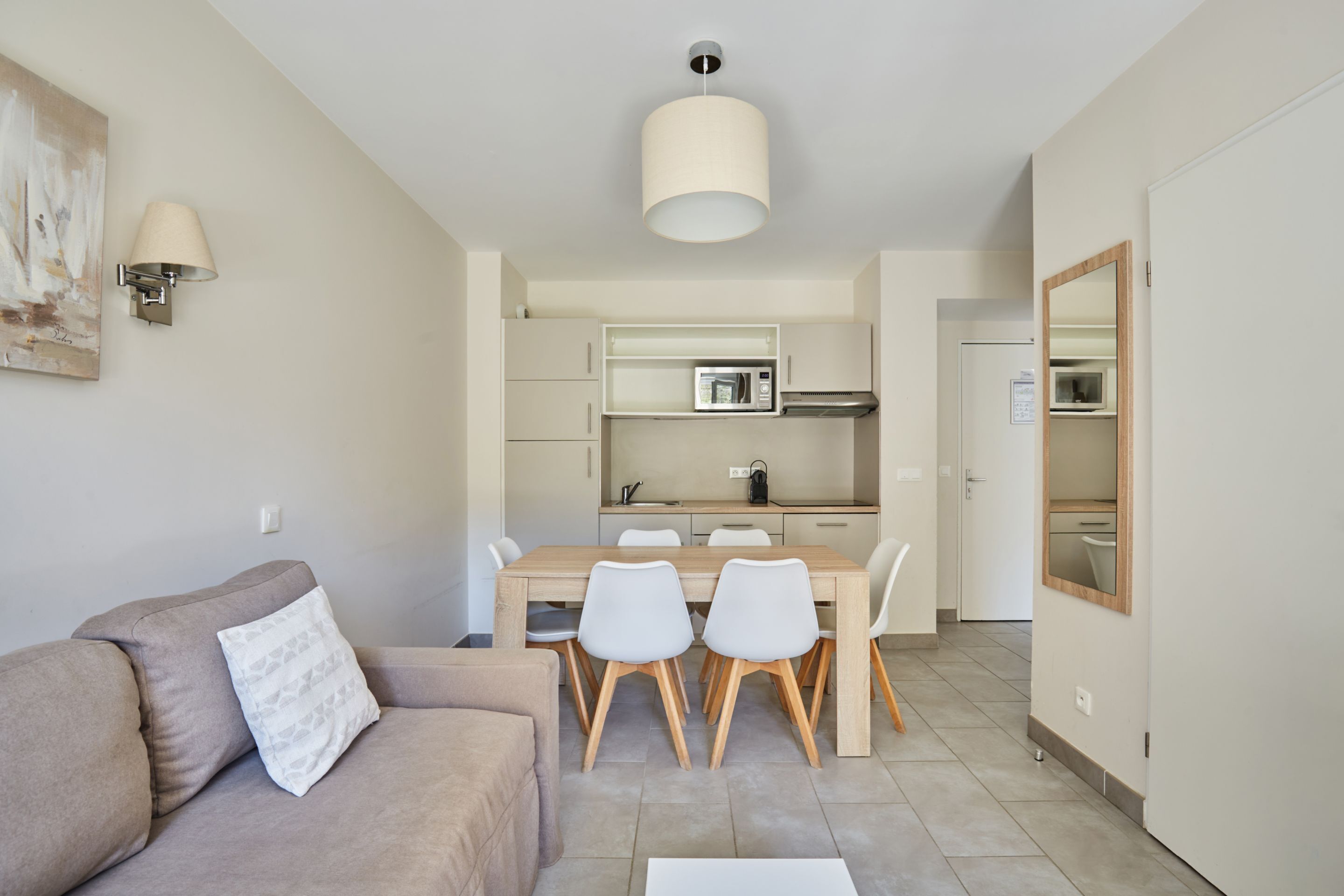 Uma sala moderna com um sofá bege, uma mesa de jantar com quatro cadeiras, uma área de cozinha com micro-ondas e um espelho grande na parede.