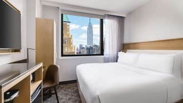 Quarto de hotel com cama, escrivaninha, cadeira, televisão e janela com vista para a cidade.