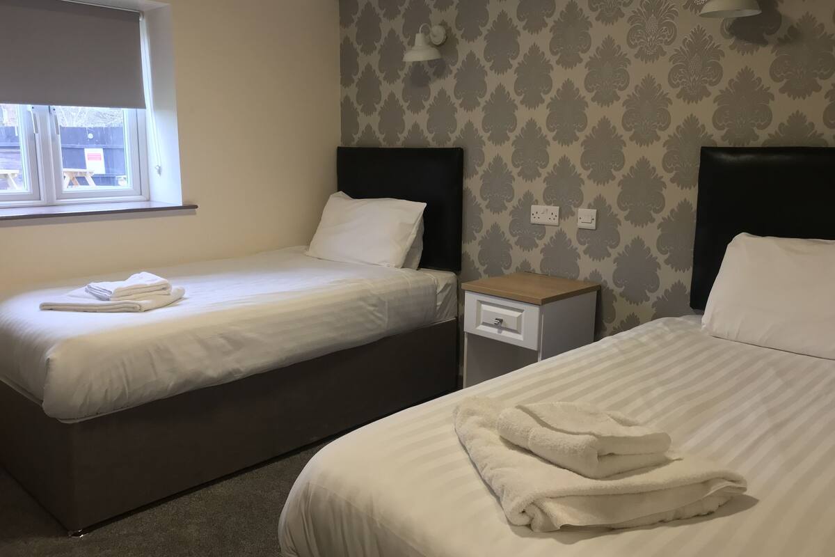 Quarto de hotel com duas camas, uma mesa de cabeceira pequena, uma janela e papel de parede estampado.