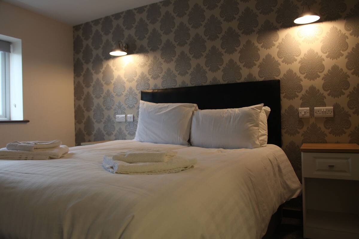 Cama bem arrumada com lençóis e toalhas brancas, cabeceira escura e papel de parede estampado.