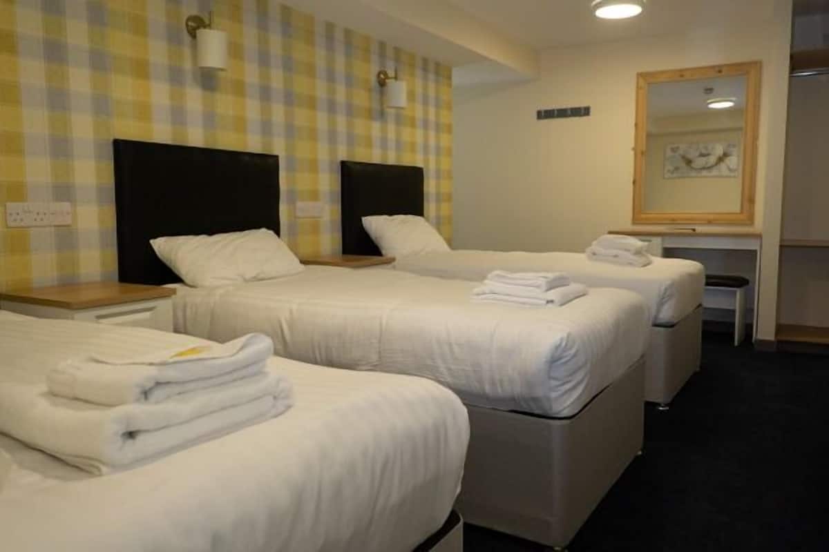 Quarto de hotel com três camas de solteiro, uma mesa com cadeira, um espelho e um relógio na parede.