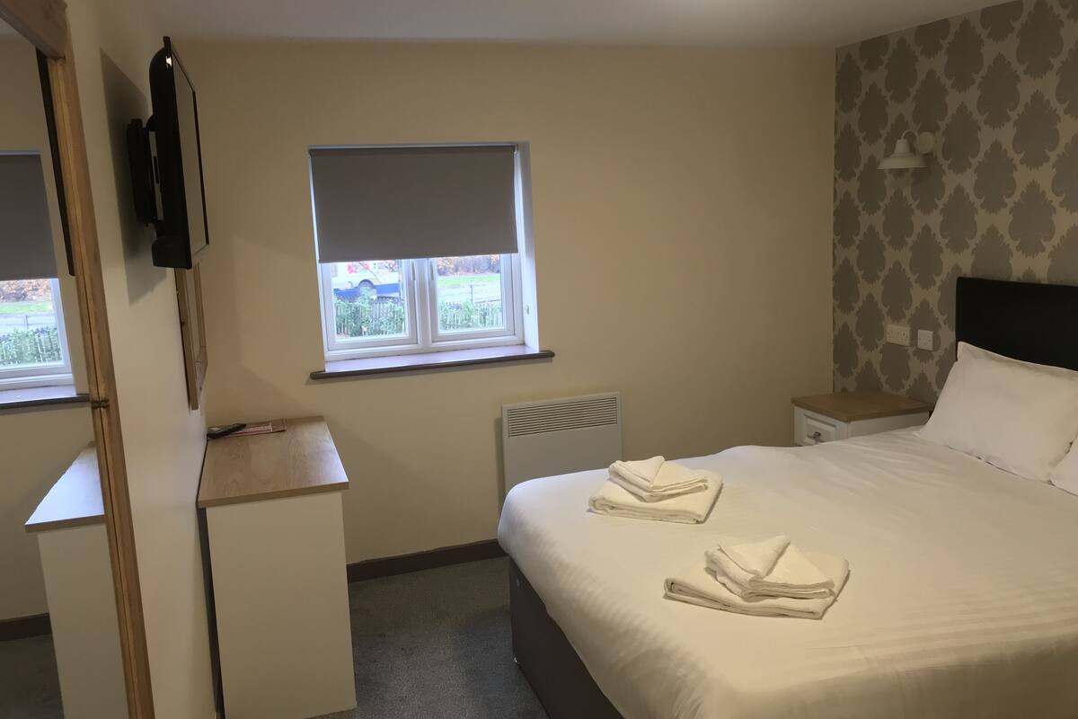 Quarto de hotel com cama, escrivaninha, janela e luminária fixada na parede.