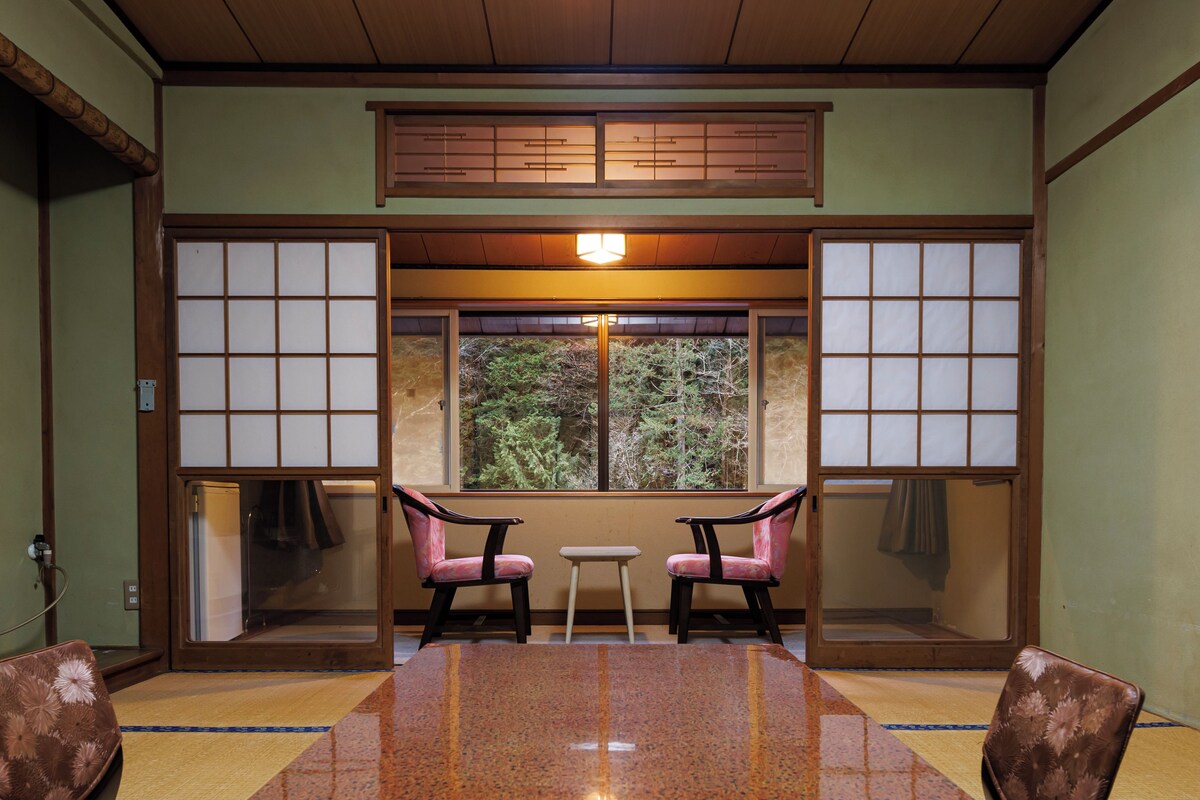 Um quarto japonês tradicional com piso de tatame, uma mesa baixa, duas cadeiras e uma janela com vista para vegetação.