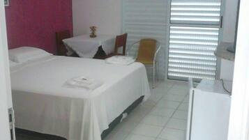 Quarto de hotel com cama, mesa pequena com cadeira, janela com persianas e parede branca.