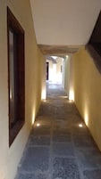 Corredor com soalho de pedra e uma janela de madeira à esquerda, que dá para um espaço interior luminoso.