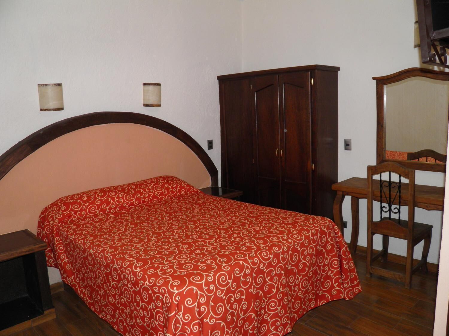 Um quarto com uma cama, um guarda-roupa de madeira, uma escrivaninha com cadeira e um espelho.