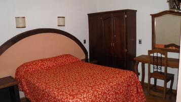 Um quarto com uma cama, um guarda-roupa de madeira, uma escrivaninha com cadeira e um espelho.