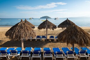 Uma praia com guarda-sóis de palha e espreguiçadeiras azuis.