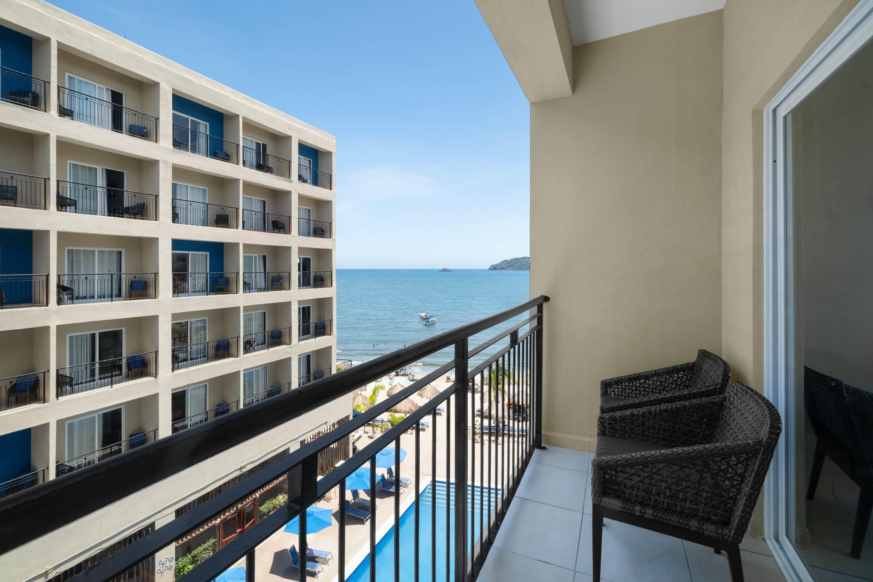 Varanda com vista para uma piscina e praia, com vista para um edifício de hotel com vários andares e varandas.