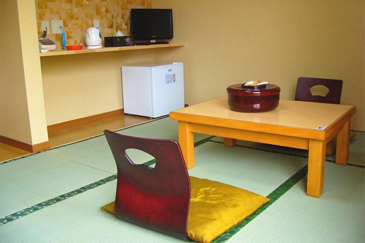 Quarto com piso de tatame, mesa de madeira, televisão em uma prateleira, geladeira e cadeira com encosto recortado.
