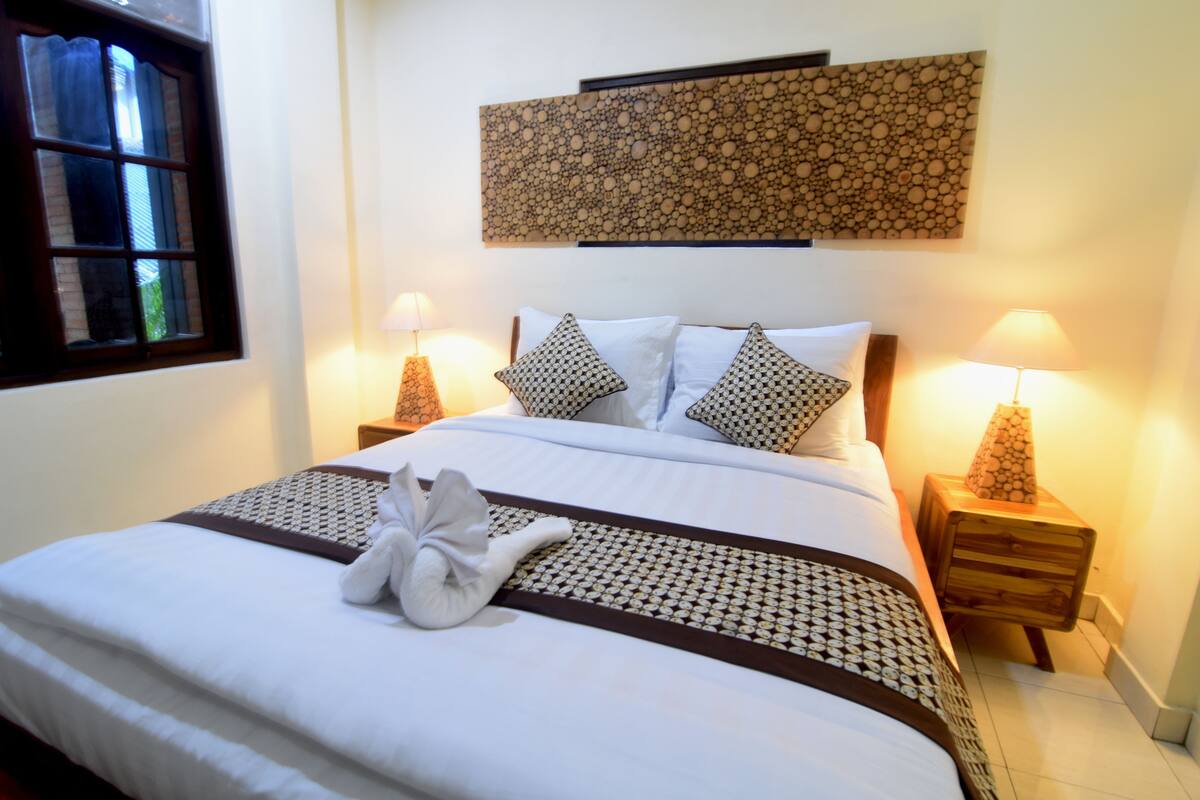 Cama bem arrumada com colcha estampada, dois travesseiros com design geométrico, um objeto decorativo sobre a cama e duas luminárias de cabeceira em mesas de cabeceira de madeira.