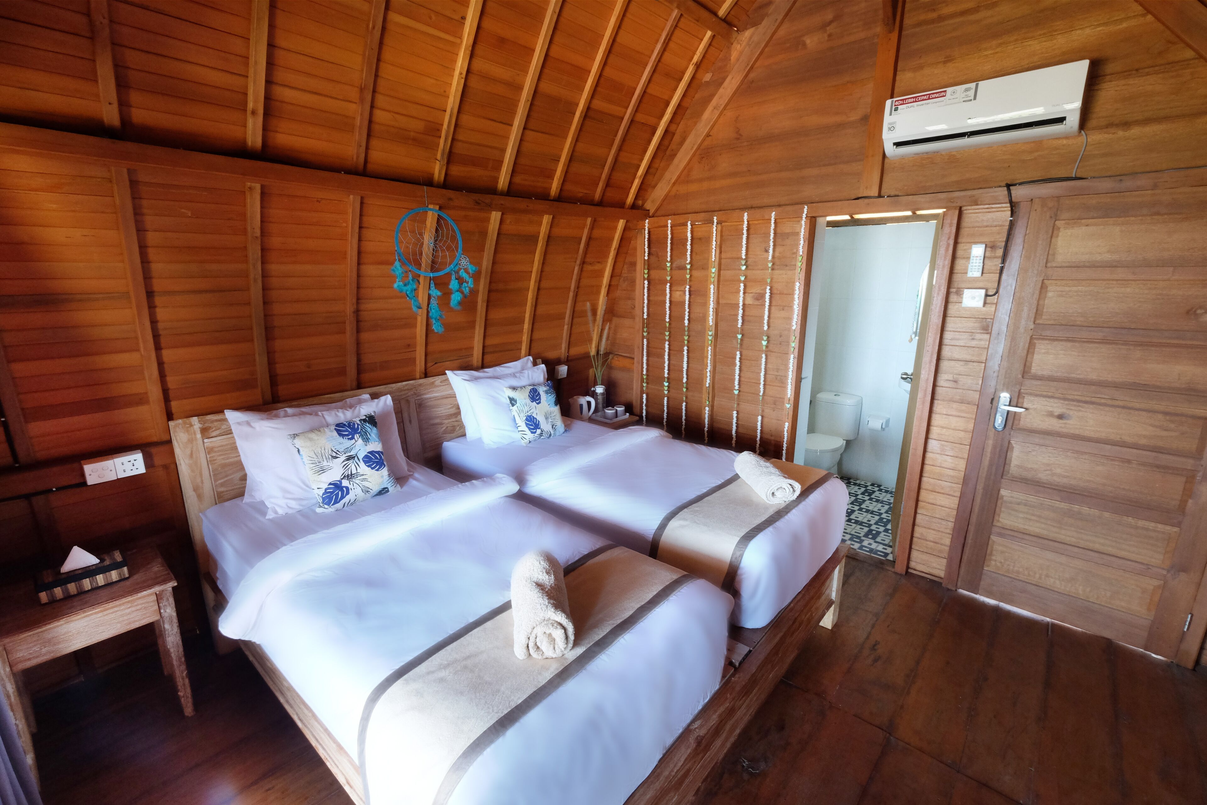 Quarto de cabana de madeira com duas camas, uma mesinha de cabeceira, um apanhador de sonhos e um aparelho de ar condicionado.