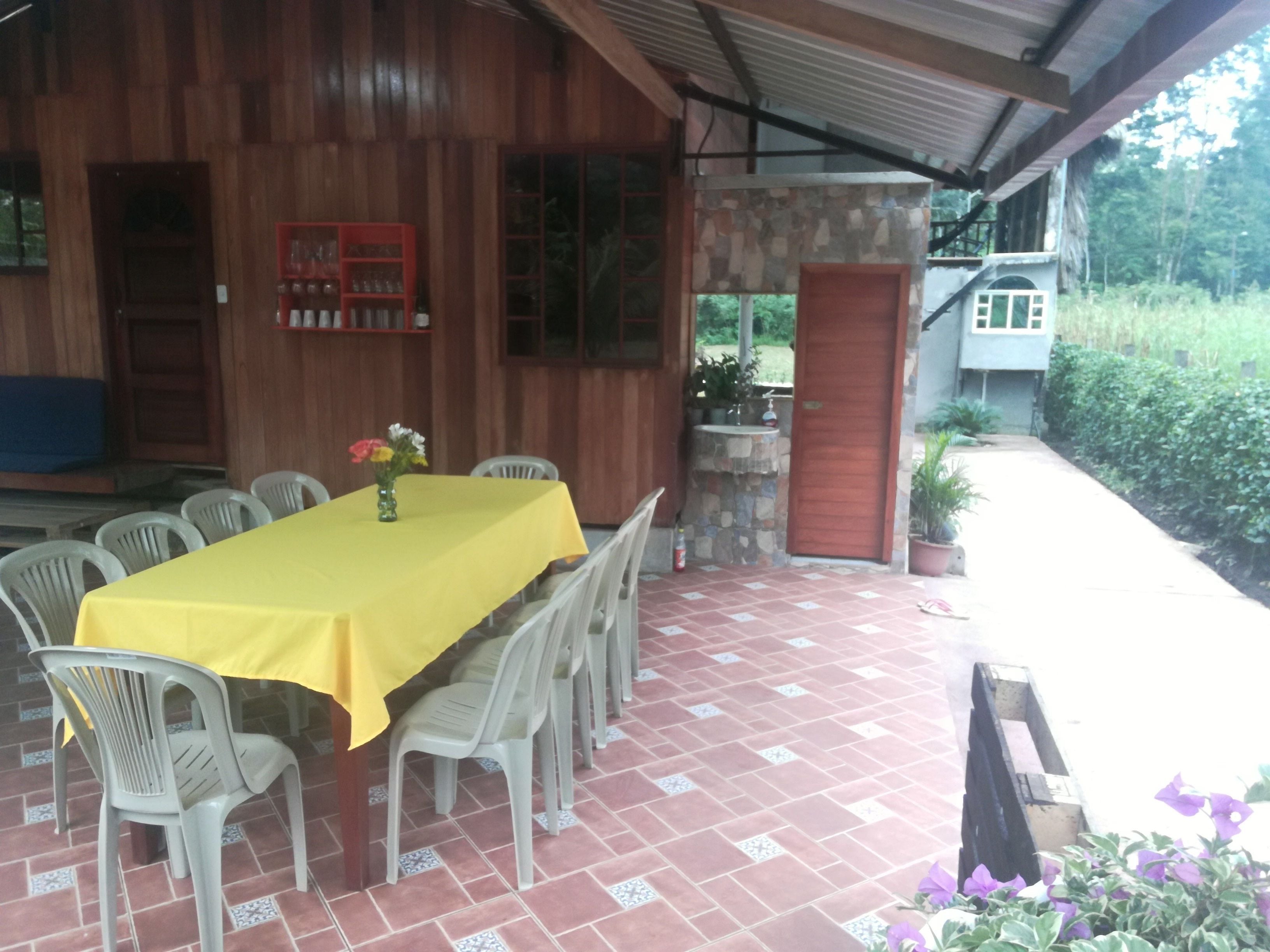Uma área de refeições ao ar livre coberta, com uma mesa posta para seis pessoas, cadeiras ao redor e uma parede de pedra com uma porta.