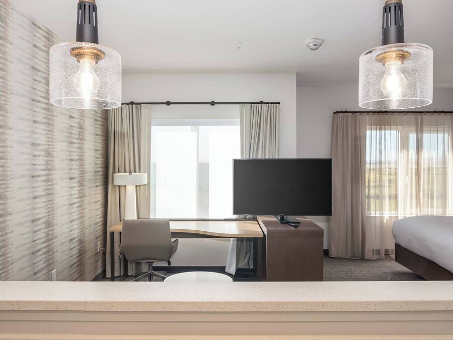 Quarto de hotel com cama, escrivaninha, cadeira, televisão e duas luzes pendentes.