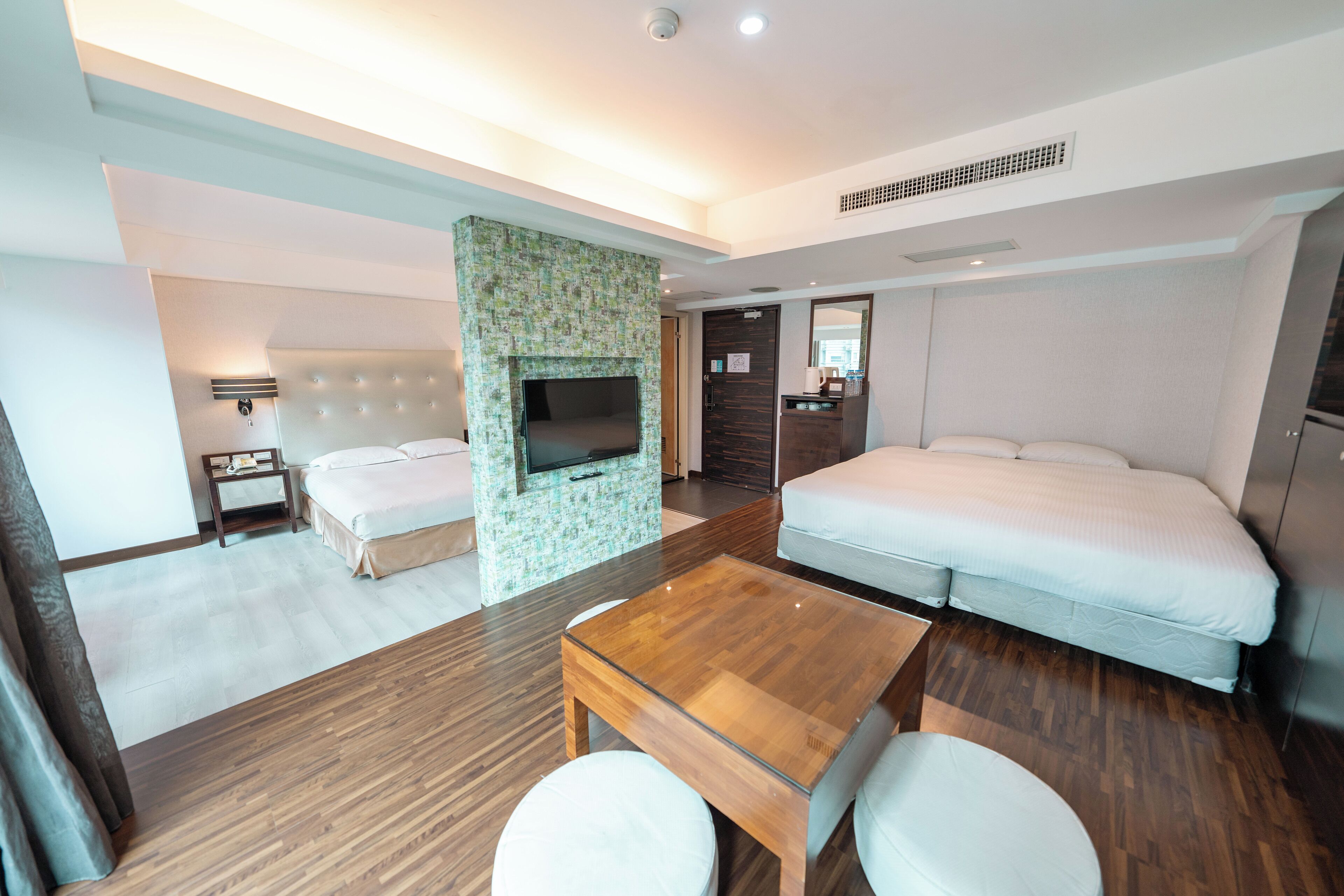 Quarto de hotel moderno com piso de madeira, cama grande, TV fixada em uma parede texturizada, mesa pequena e área de estar.