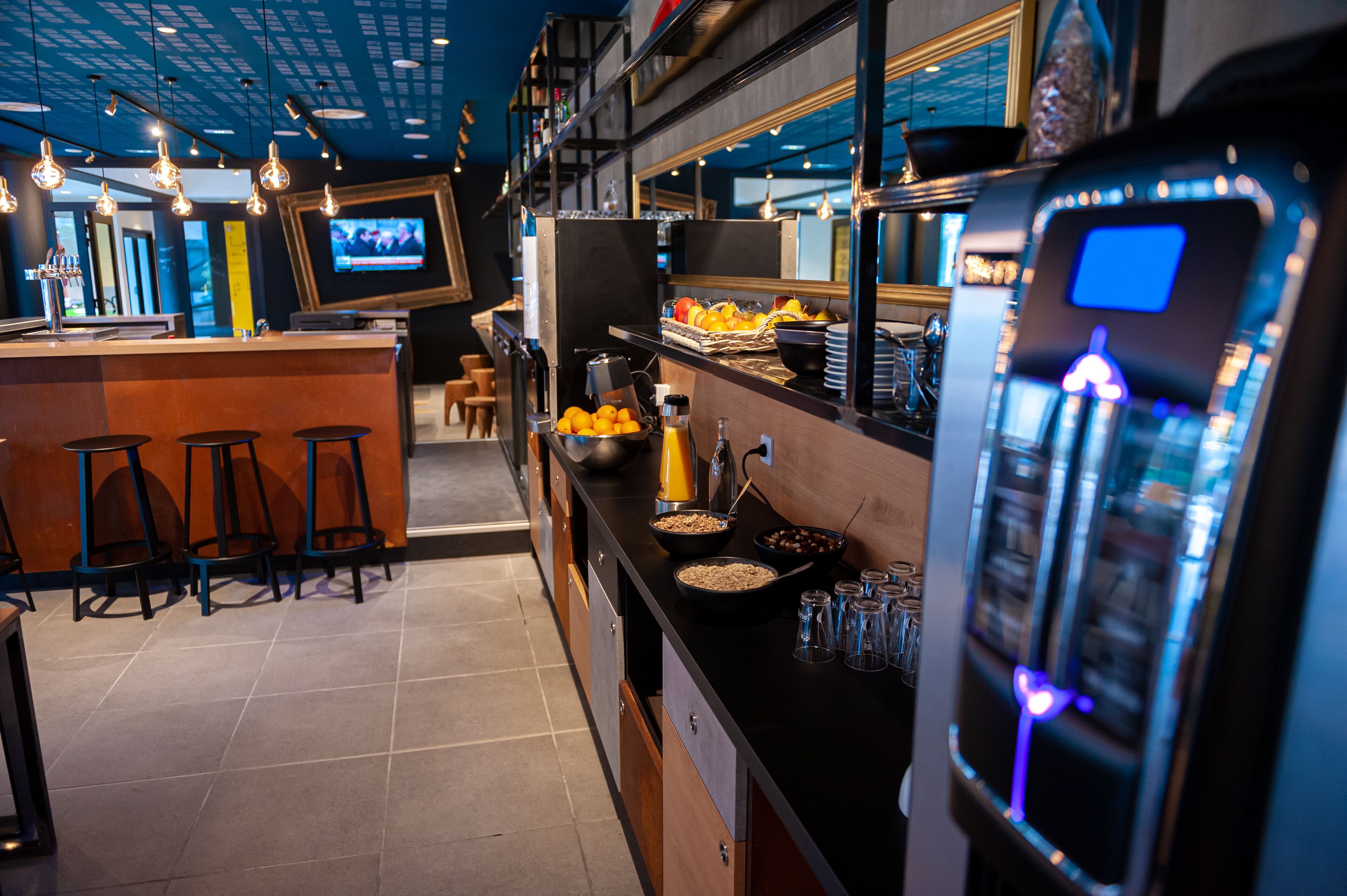 Uma área de bar moderna, com banquetas, um balcão com opções de comida e uma tela de televisão.