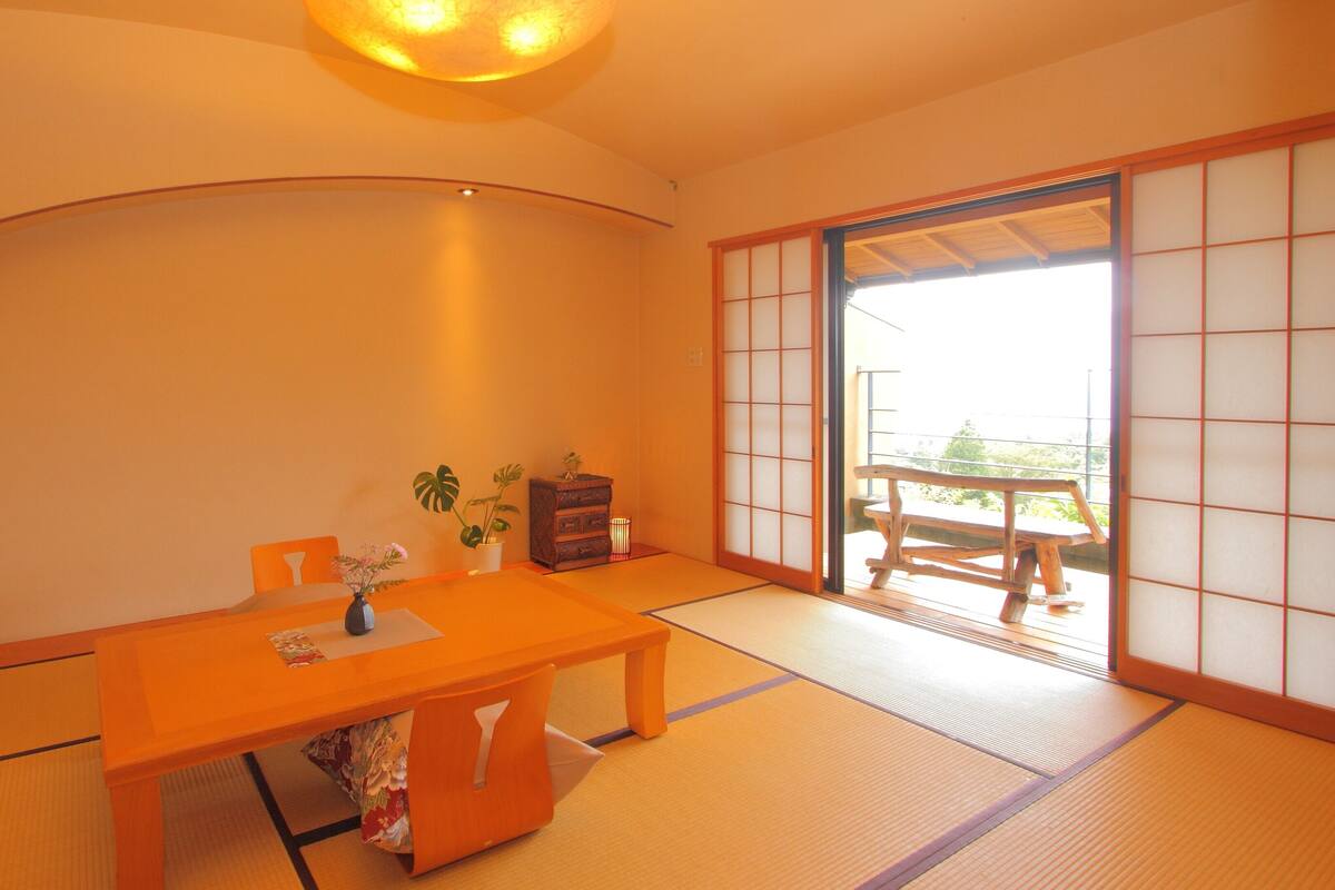 Um quarto japonês tradicional com tatames, uma mesa baixa, uma cadeira, um pequeno armário e uma porta deslizante que dá para uma área externa.