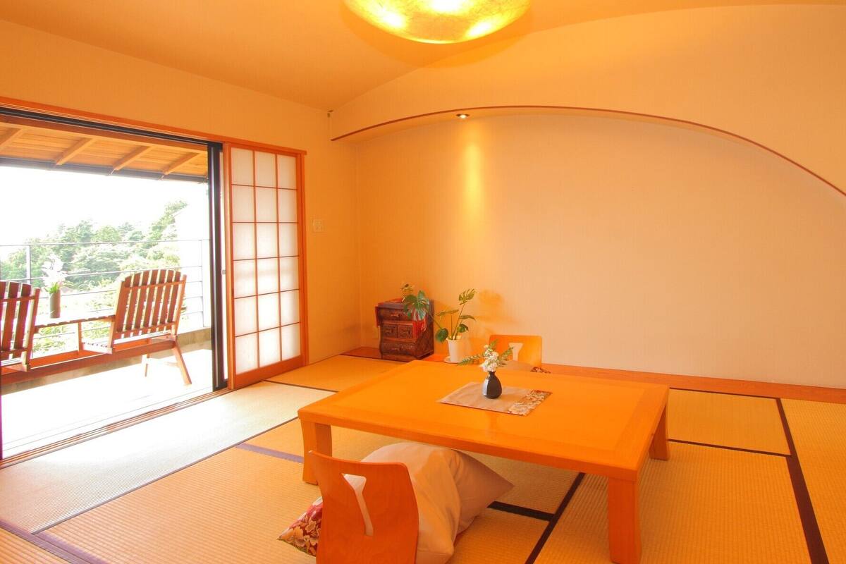 Quarto com piso de tatame, uma mesa baixa, uma cadeira, um pequeno armário e uma porta deslizante que dá para uma varanda.