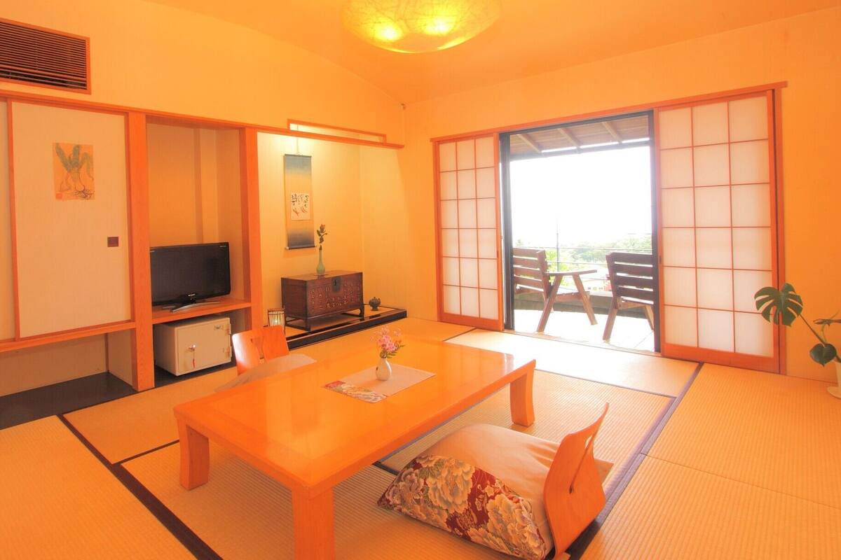 Um quarto japonês tradicional com tatames, televisão, armário e uma porta deslizante que dá para uma varanda.