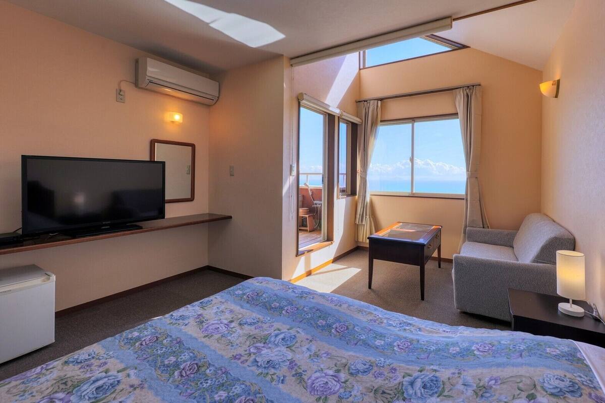 Quarto de hotel com cama, televisão, escrivaninha, cadeira e varanda com vista.