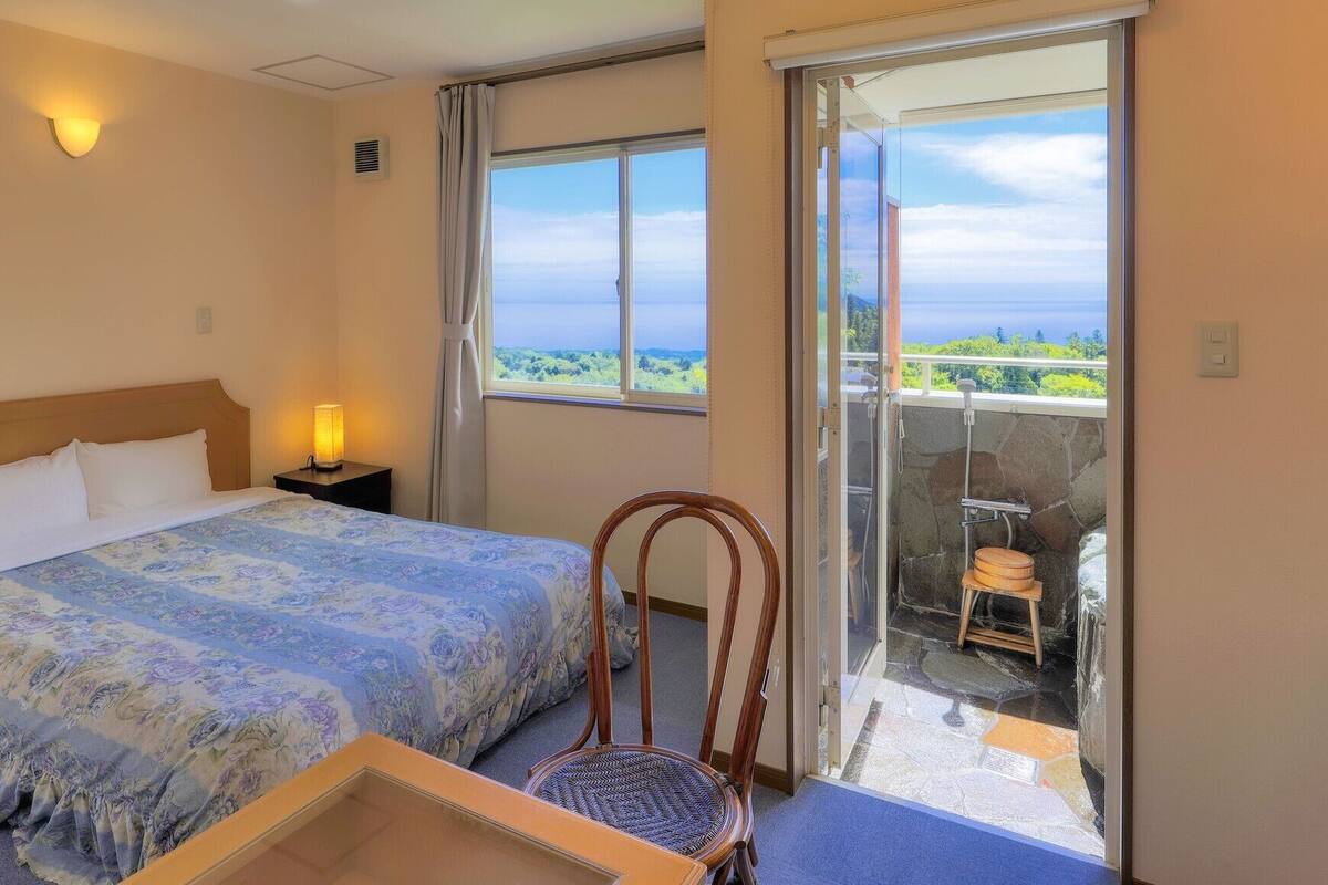 Quarto com cama, mesa de cabeceira, abajur, cadeira e varanda com vista.