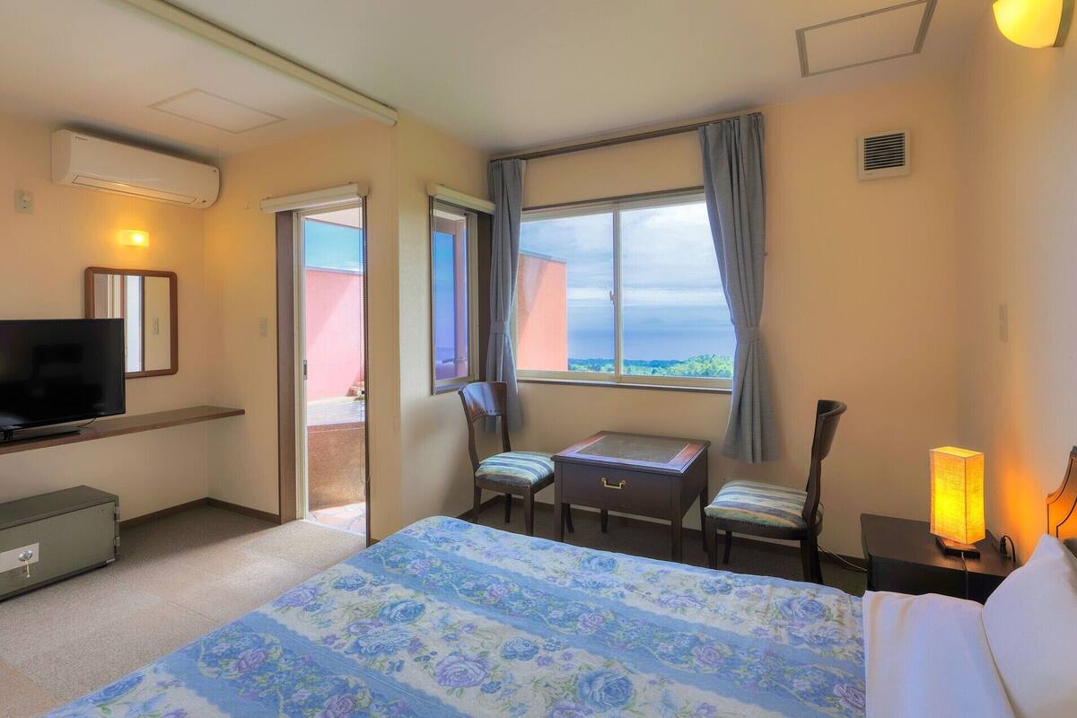 Quarto de hotel com cama, escrivaninha, cadeira, televisão, cofre e janela com vista.