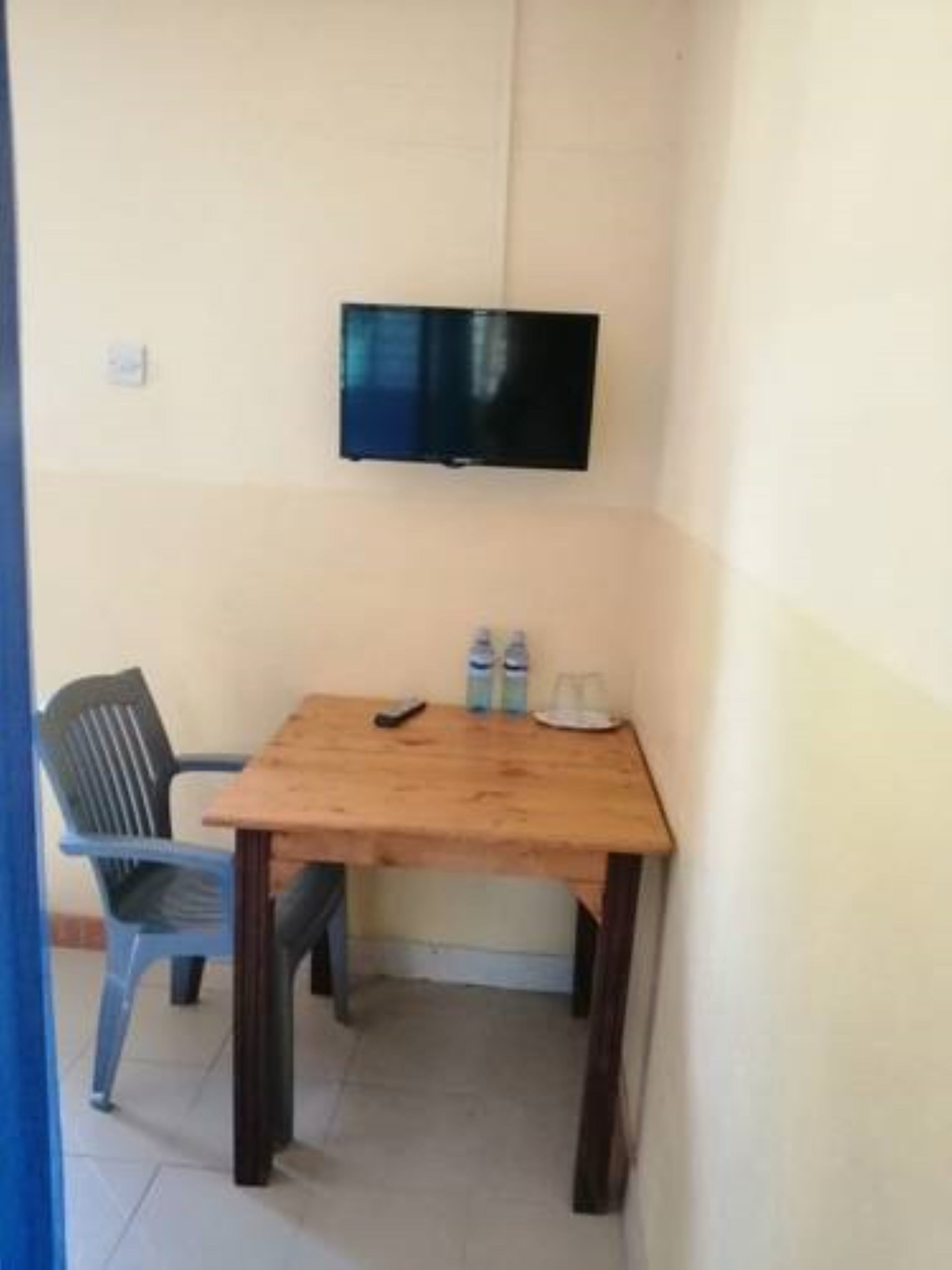 Um quarto pequeno com uma mesa de madeira, uma cadeira azul, uma televisão fixada na parede e algumas garrafas sobre a mesa.