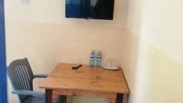 Um quarto pequeno com uma mesa de madeira, uma cadeira azul, uma televisão fixada na parede e algumas garrafas sobre a mesa.