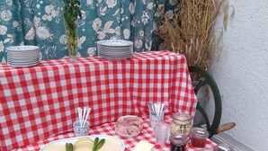 Mesa posta com diversos alimentos e utensílios, emoldurada por cortinas florais e um cenário rústico.