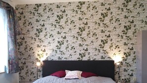 Cama bem arrumada com colcha acolchoada, dois travesseiros e duas toalhas dobradas. O quarto tem parede com padrão de tijolos e trepadeiras verdes, e duas luminárias de cabeceira.