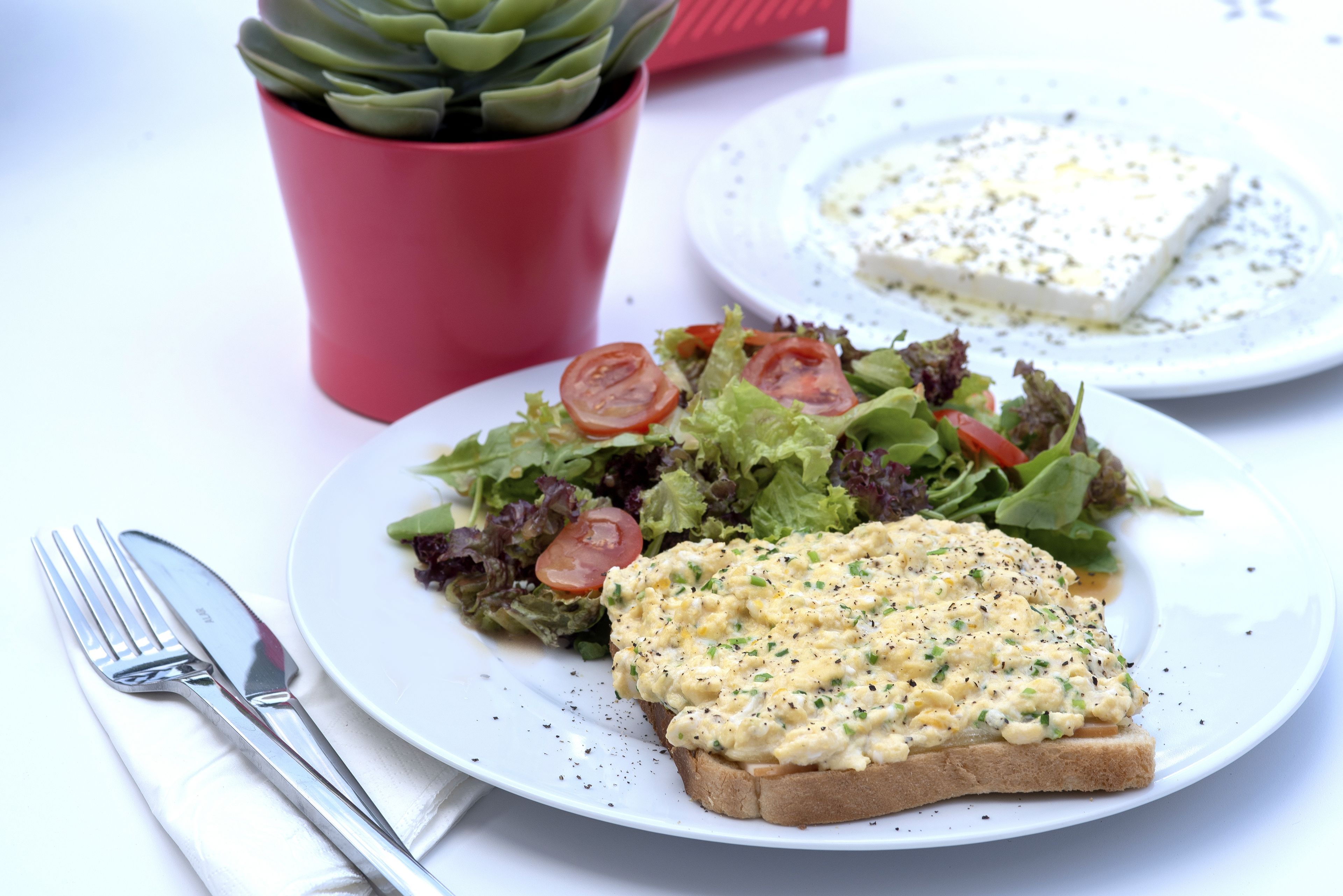 Prato com salada e uma fatia de pão torrado coberta por um creme, acompanhado de garfo e faca sobre uma toalha de mesa branca.