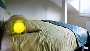 Um quarto com cama, luminária, janela e um quadro na parede.